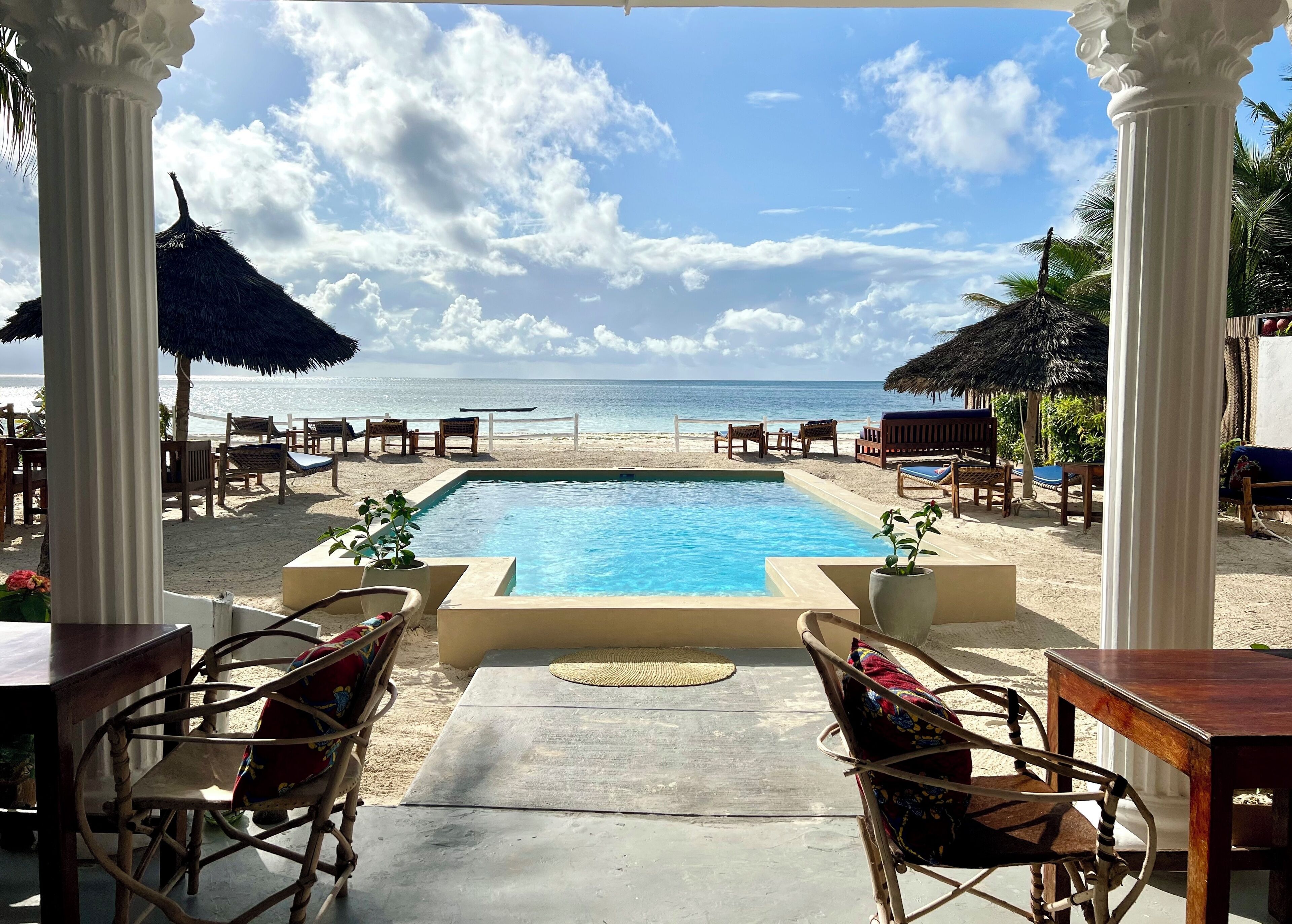 Área da piscina com vista para a praia e oceano, cadeiras de madeira e guarda-sóis de palha.