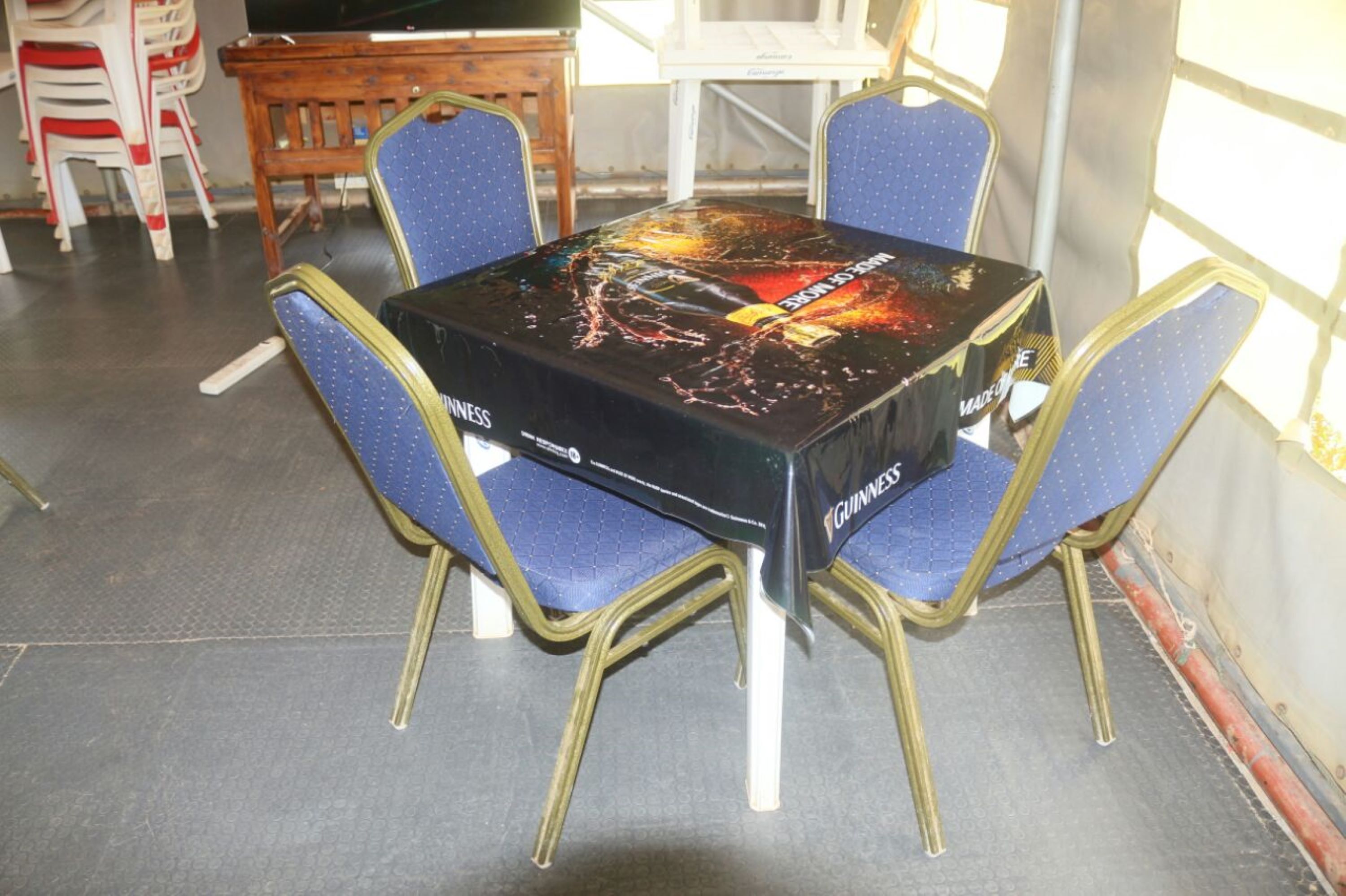 Uma mesa com uma caixa de cerveja Guinness, cadeiras azuis e uma mesa de madeira ao fundo.