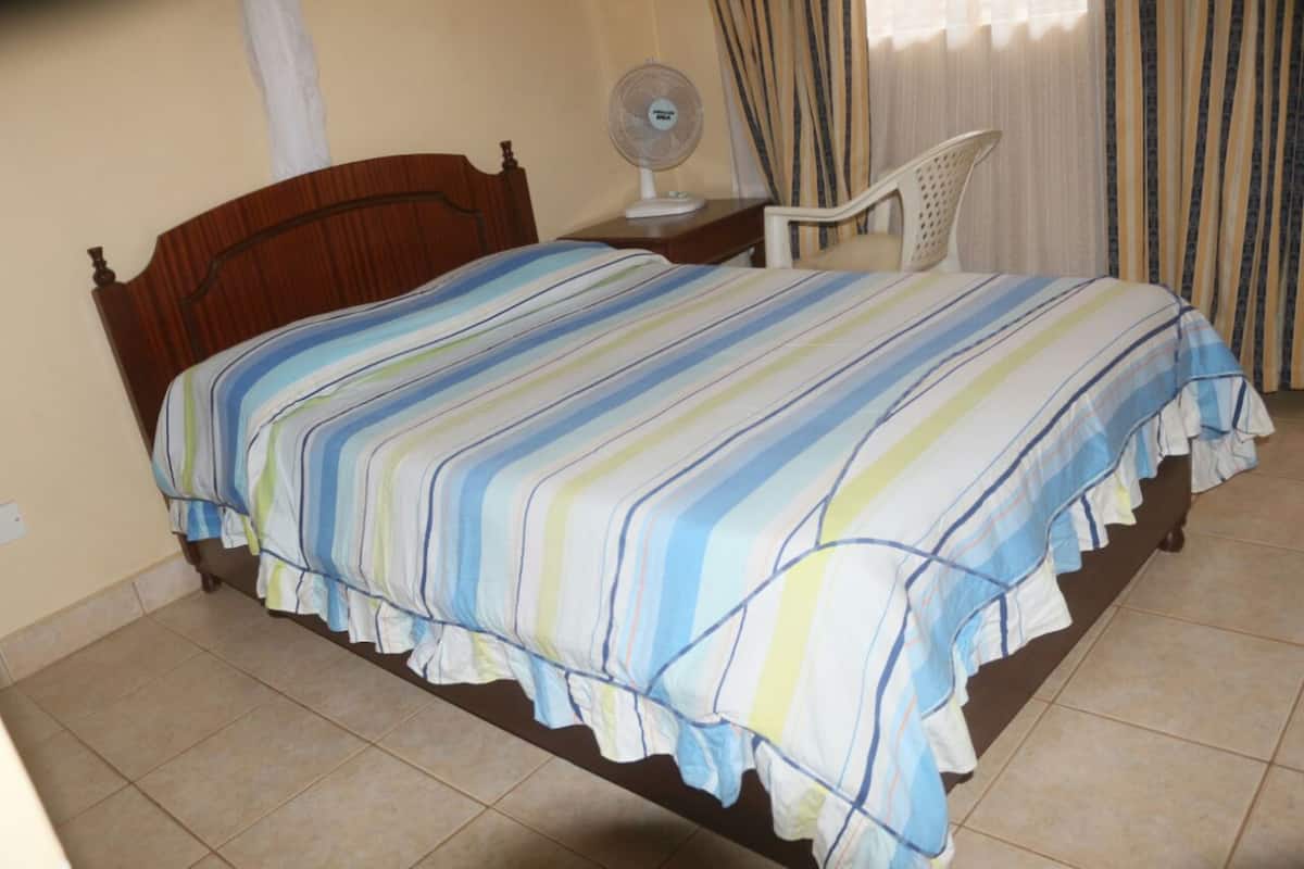 Uma cama com colcha listrada, uma mesa de cabeceira com um ventilador e uma cadeira.