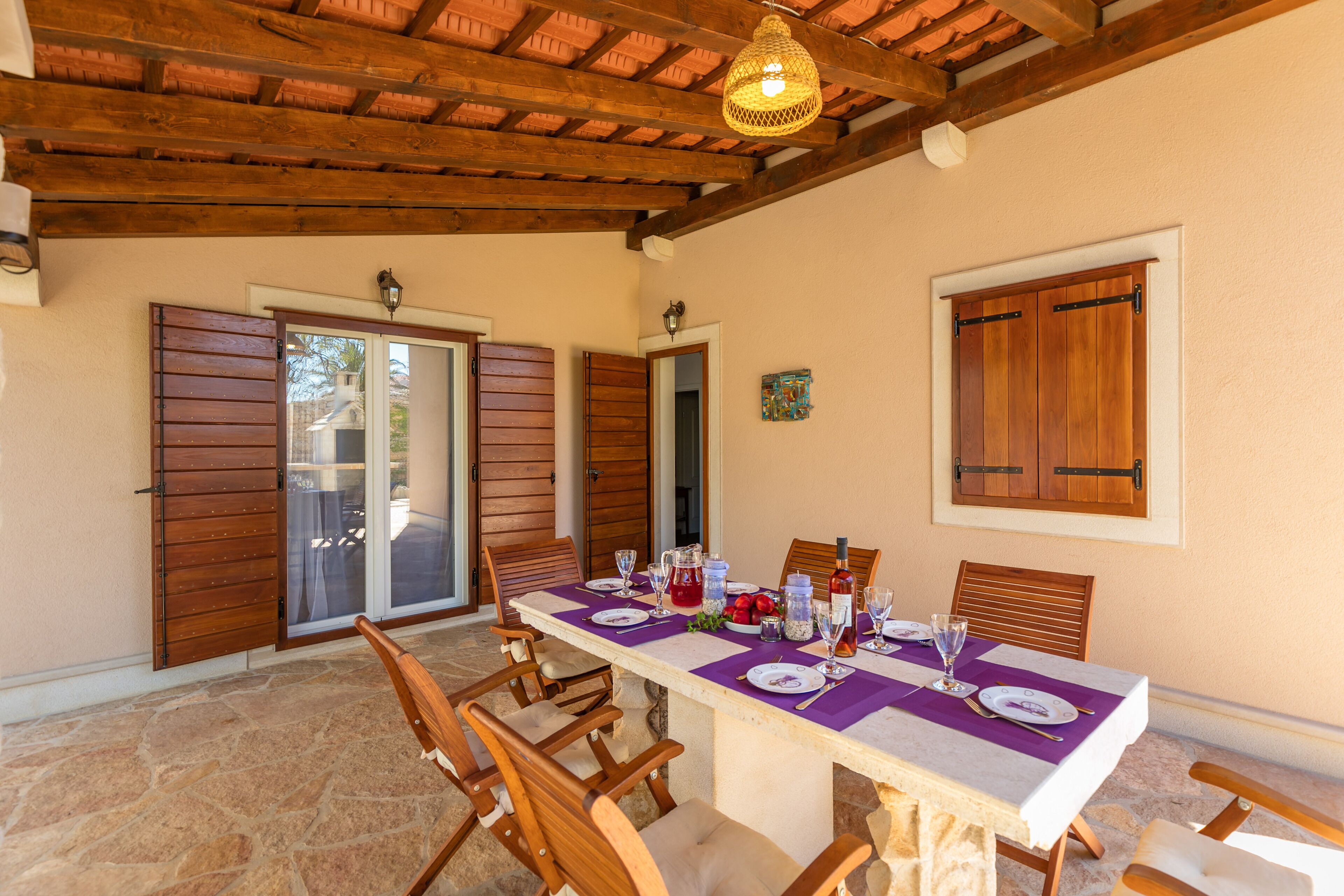 Uma sala de jantar com piso de pedra, vigas de madeira no teto e uma mesa posta para quatro pessoas, com jogos americanos roxos.