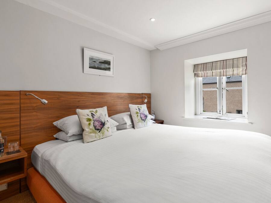 Cama bem arrumada com almofadas florais, cabeceira de madeira, mesa de cabeceira com garrafa e copo, e janela com valance listrado.