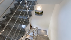 Uma escadaria com degraus de granito e guarda-corpos de metal, que leva a um patamar com um quadro fixado na parede e um candeeiro.