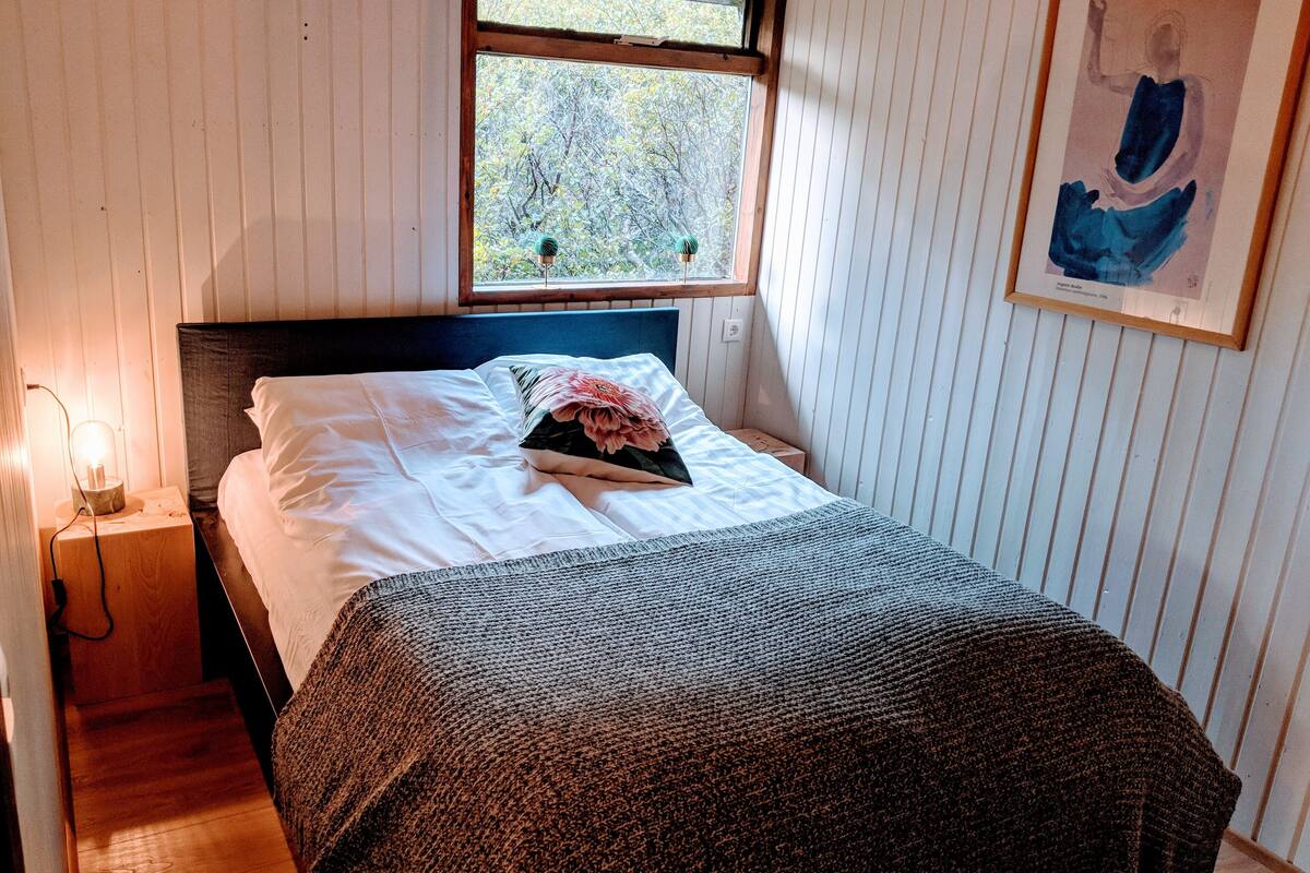 Cama bem arrumada com travesseiro floral, luminária de cabeceira e uma obra de arte emoldurada na parede.