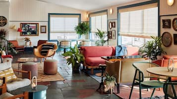 Um ambiente acolhedor com diferentes áreas de estar, incluindo um sofá vermelho, cadeiras de vime e uma mesa de jantar. Várias plantas em vasos trazem verde ao espaço, que se destaca pelas diversas texturas e padrões nas paredes e no chão.