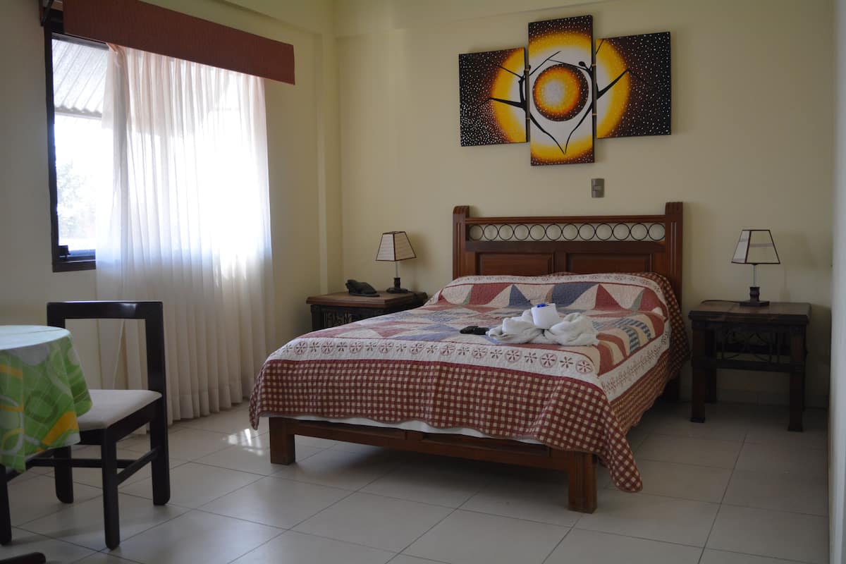 Quarto bem arrumado com cama, duas mesas de cabeceira, uma cadeira, uma mesa e uma janela com cortinas.