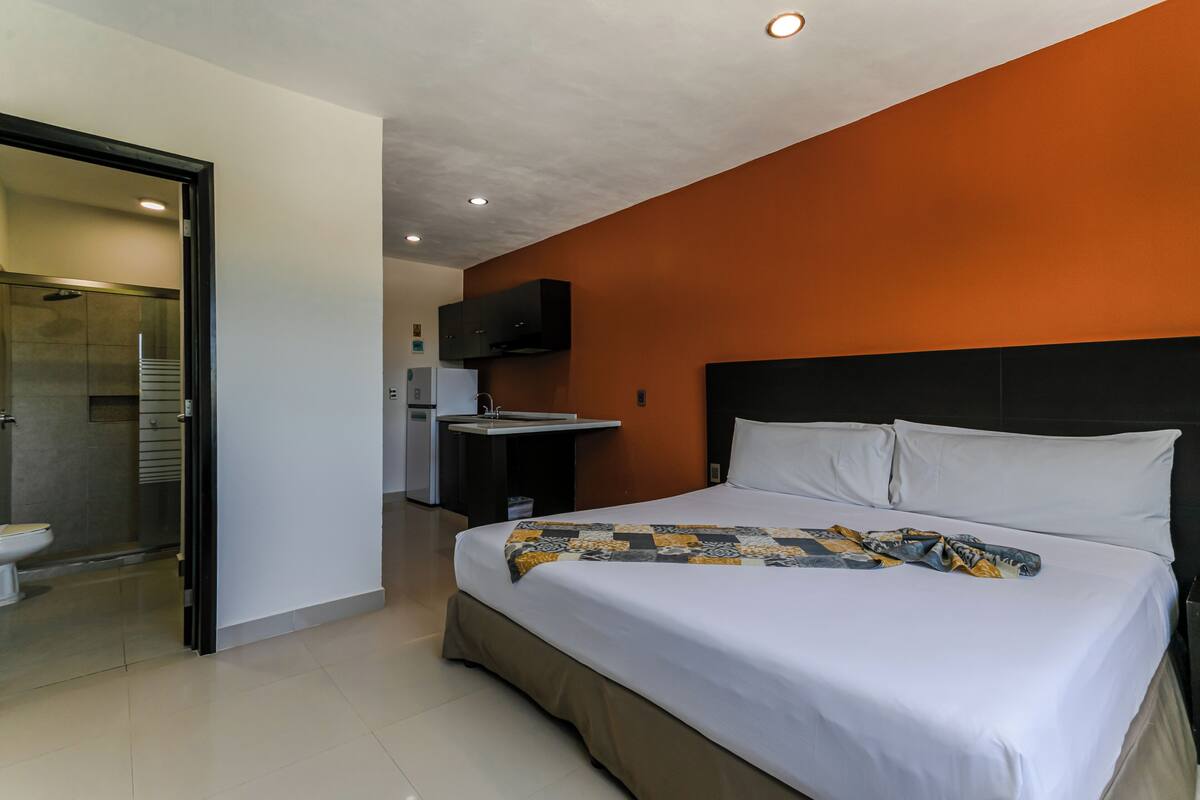 Quarto de hotel com cama, banheiro com chuveiro e cozinha compacta.