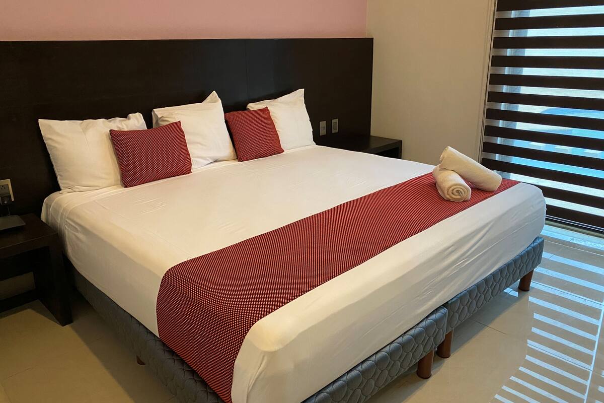 Uma cama bem arrumada com roupa de cama branca e vermelha, cabeceira escura e uma janela com persianas.