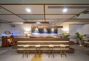 Uma área de recepção com balcão de madeira, três banquetas, um quadro decorativo grande, um pequeno santuário com flores e uma planta em vaso.