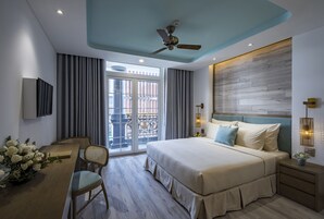 Quarto de hotel moderno com uma cama grande, uma escrivaninha com cadeira, uma varanda com cortinas e um ventilador de teto.