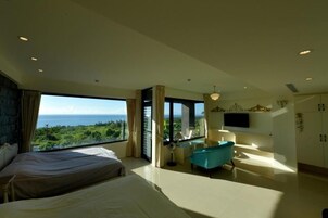 Quarto de hotel moderno com uma cama grande, um espaço de estar com uma cadeira azul, uma TV fixada na parede e vista para o oceano.
