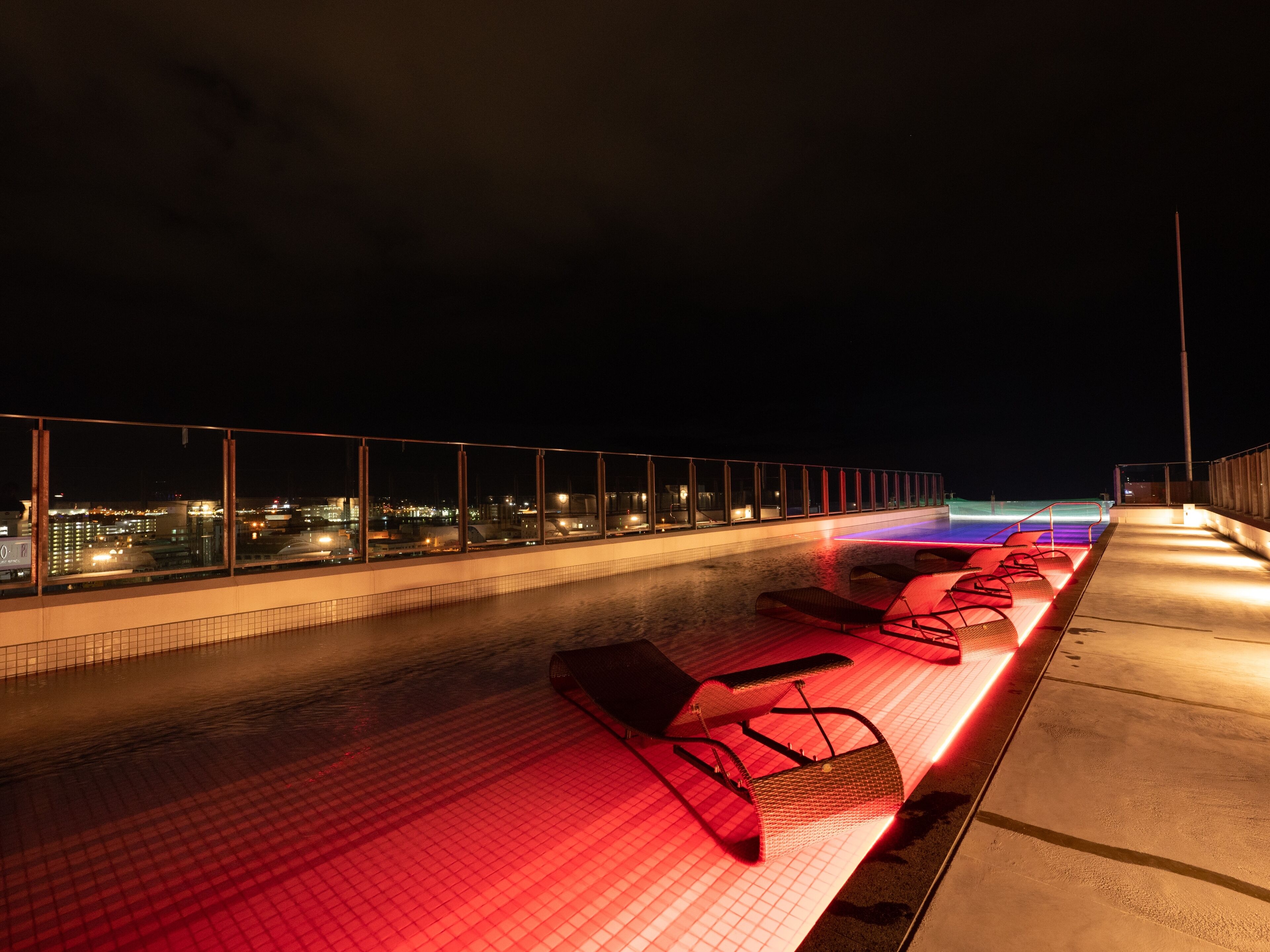 Área de piscina no terraço com espreguiçadeiras e vista para a cidade à noite.
