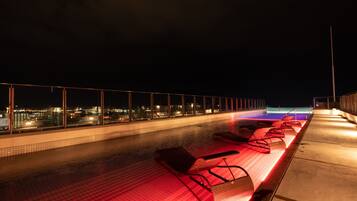 Área de piscina no terraço com espreguiçadeiras e vista para a cidade à noite.
