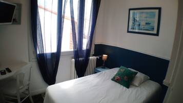 Quarto de hotel com cama, escrivaninha, cadeira, radiador, luminária e um quadro na parede.