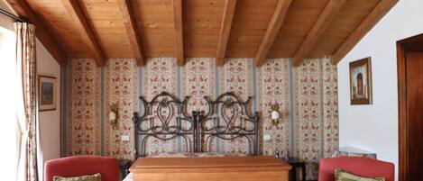 Quarto com teto de madeira, papel de parede floral, cabeceira de metal e duas poltronas com almofadas.