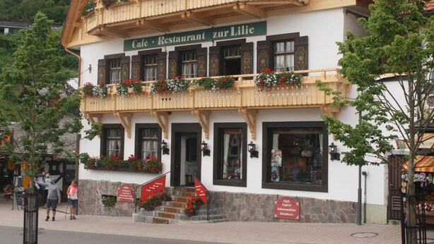 Um edifício tradicional com um café e restaurante chamados 'zur Lilie'. Apresenta um balcão de madeira com caixas de flores, uma base de pedra e várias janelas.