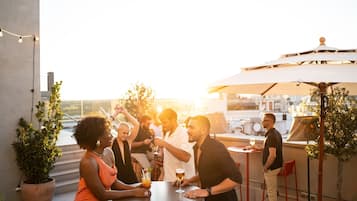 Um bar no terraço com mesas ao ar livre, plantas em vasos e vista para o horizonte da cidade.