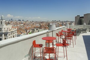 Terraço no telhado com cadeiras e mesas vermelhas, oferecendo vista para um horizonte urbano com edifícios e céu claro.