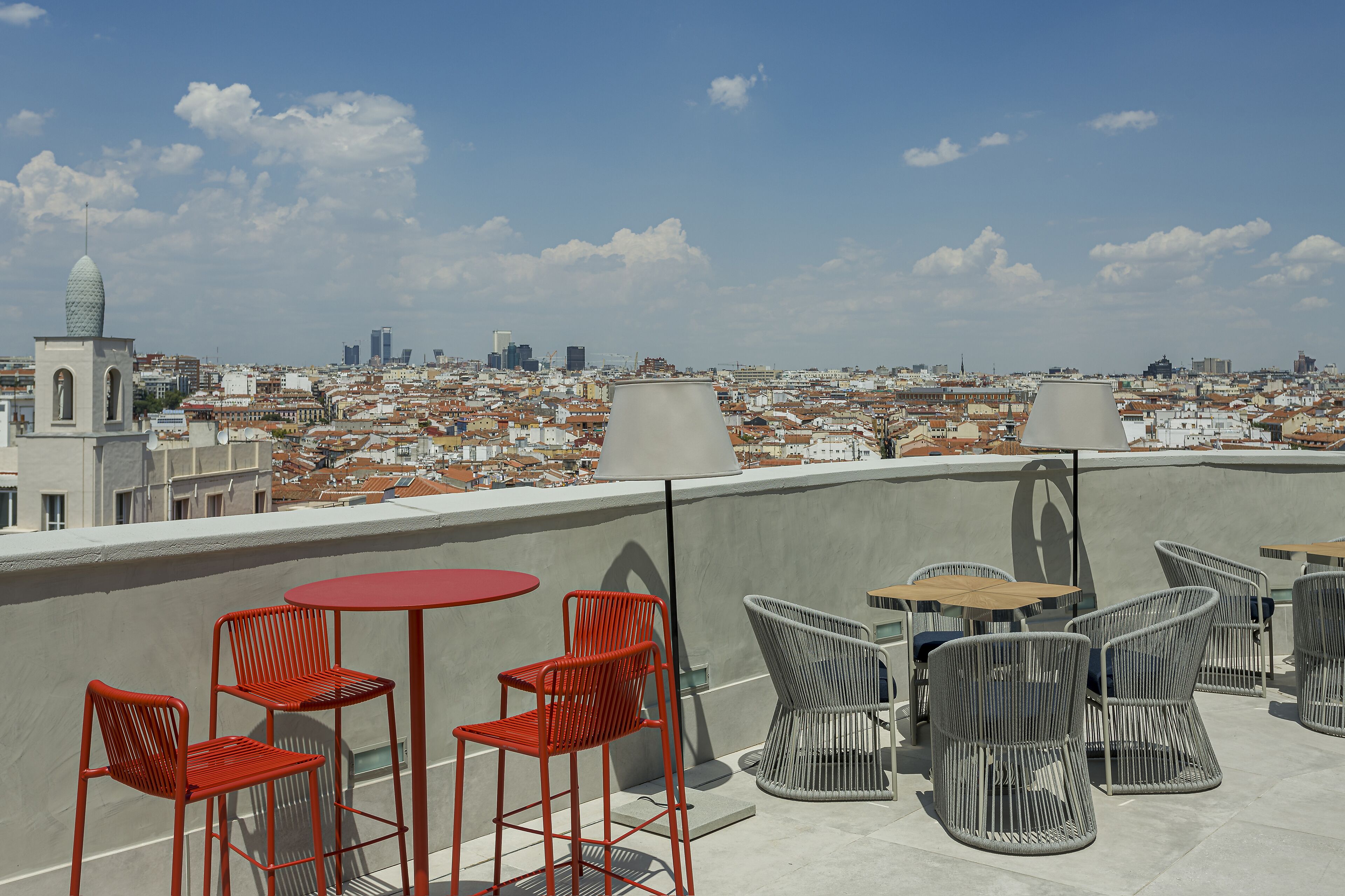 Um terraço no telhado com cadeiras de vime vermelhas e cinzentas, uma mesa vermelha e vista para a paisagem urbana.