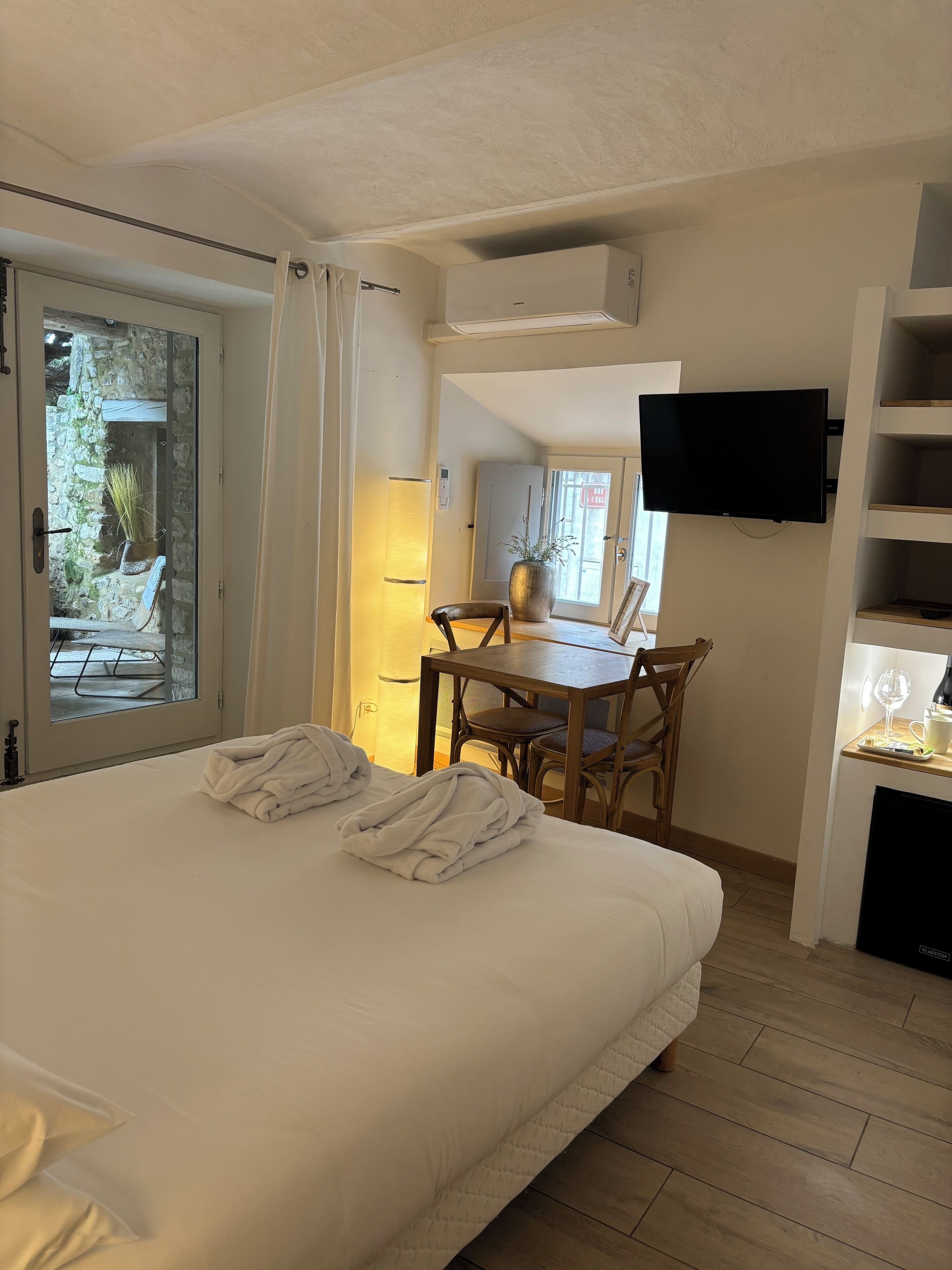 Quarto de hotel com uma cama grande, uma mesa de jantar com cadeiras, uma televisão fixada na parede e uma pequena cozinha.
