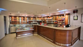 Uma área de bar bem iluminada e moderna, com bancada de mármore, prateleiras cheias de petiscos e bebidas, e uma vitrine de vidro.