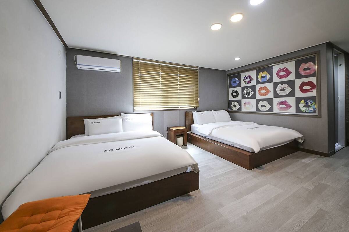 Quarto de hotel com duas camas, janela com persianas, ar condicionado fixado na parede e uma parede com obras de arte emolduradas.