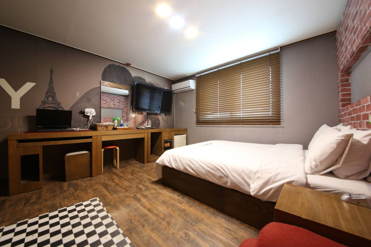 Quarto com cama, escrivaninha com computador, televisão e janela com persianas.