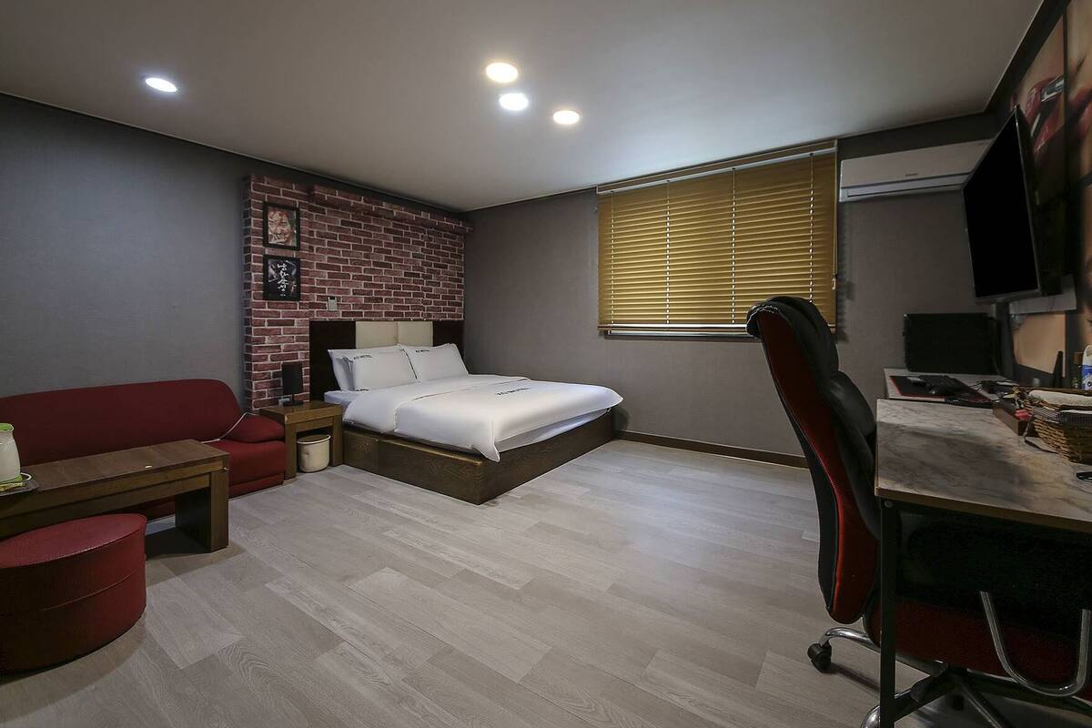 Um quarto moderno com parede de tijolos, uma cama, um sofá vermelho, uma mesa de trabalho com cadeira e uma janela com persianas.