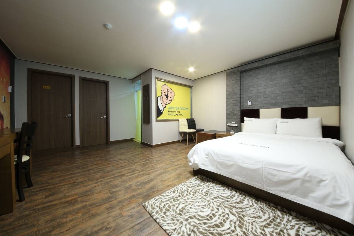 Quarto de hotel moderno com uma cama grande, uma mesa e uma cadeira. Há duas portas e um quadro na parede.