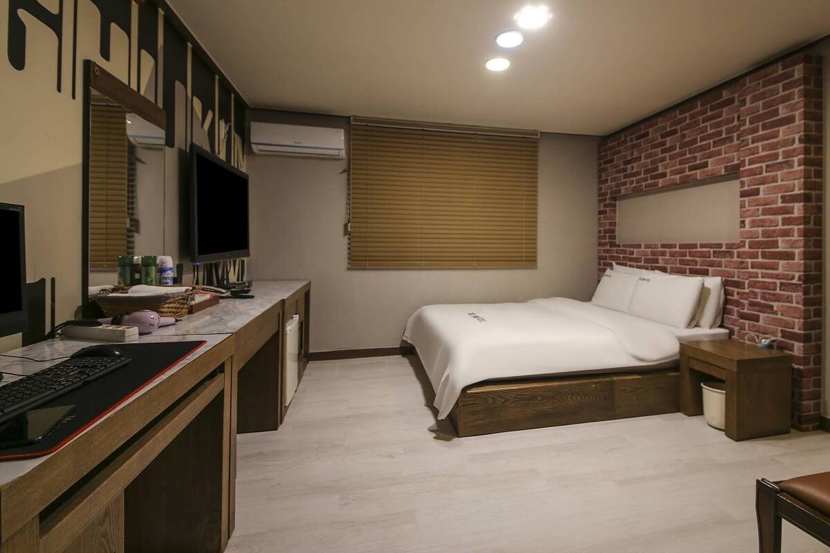 Quarto com escrivaninha, cama, parede de tijolos e janela com persianas.