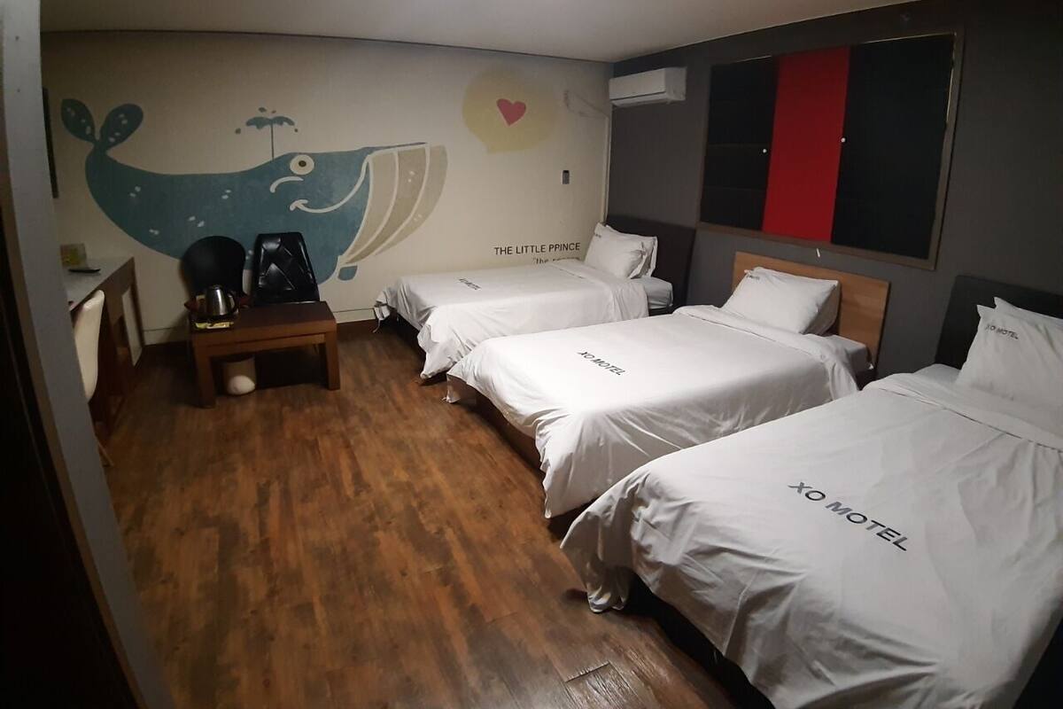 Quarto de hotel com três camas, piso de madeira, mural de uma baleia na parede e painel vermelho e preto.