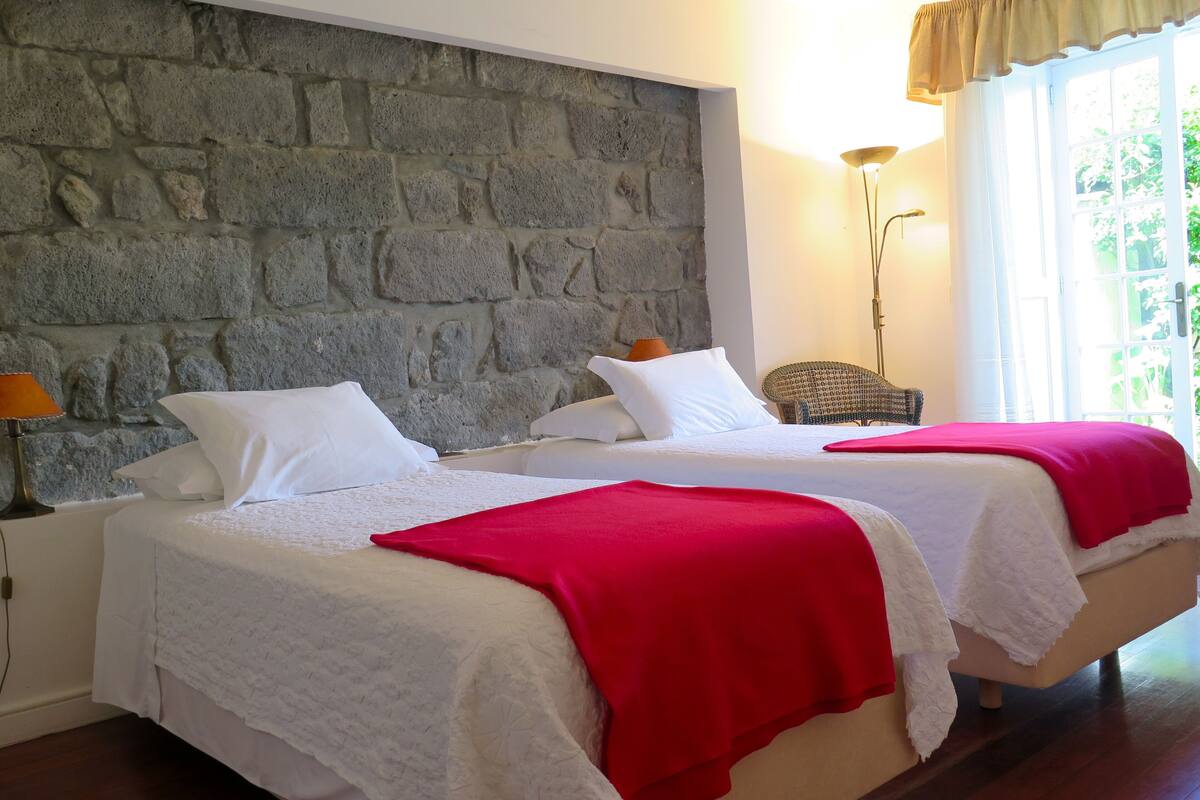 Quarto com duas camas, parede de pedra, cadeira de vime e um abajur.