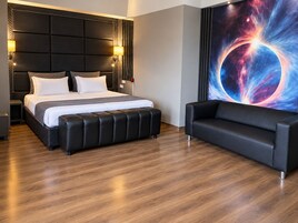 Quarto de hotel moderno com cama grande, um banquinho de couro preto, sofá e um mural na parede com cena cósmica.