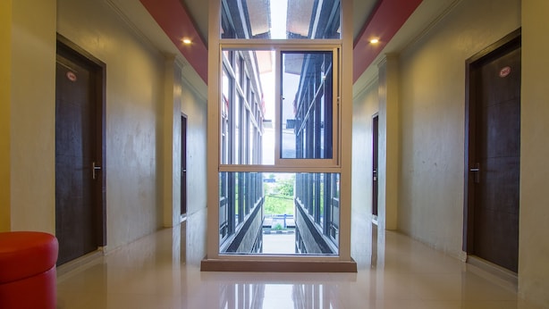 Corredor com elevador de vidro central, portas em ambos os lados e um banco vermelho em primeiro plano.