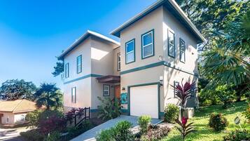 Uma casa residencial de dois andares com garagem, rodeada por vegetação exuberante e um céu azul límpido.