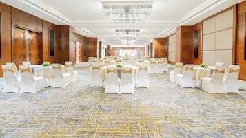 Uma ampla sala de banquetes com mesas redondas e cadeiras brancas, apresentando um carpete estampado e painéis de madeira.