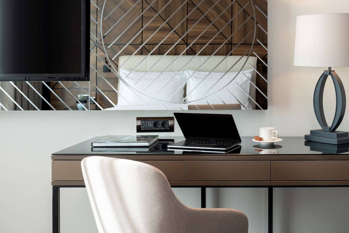 Quarto de hotel moderno com cama, escrivaninha com laptop, televisão de tela plana e mesa de cabeceira com abajur e uma xícara.