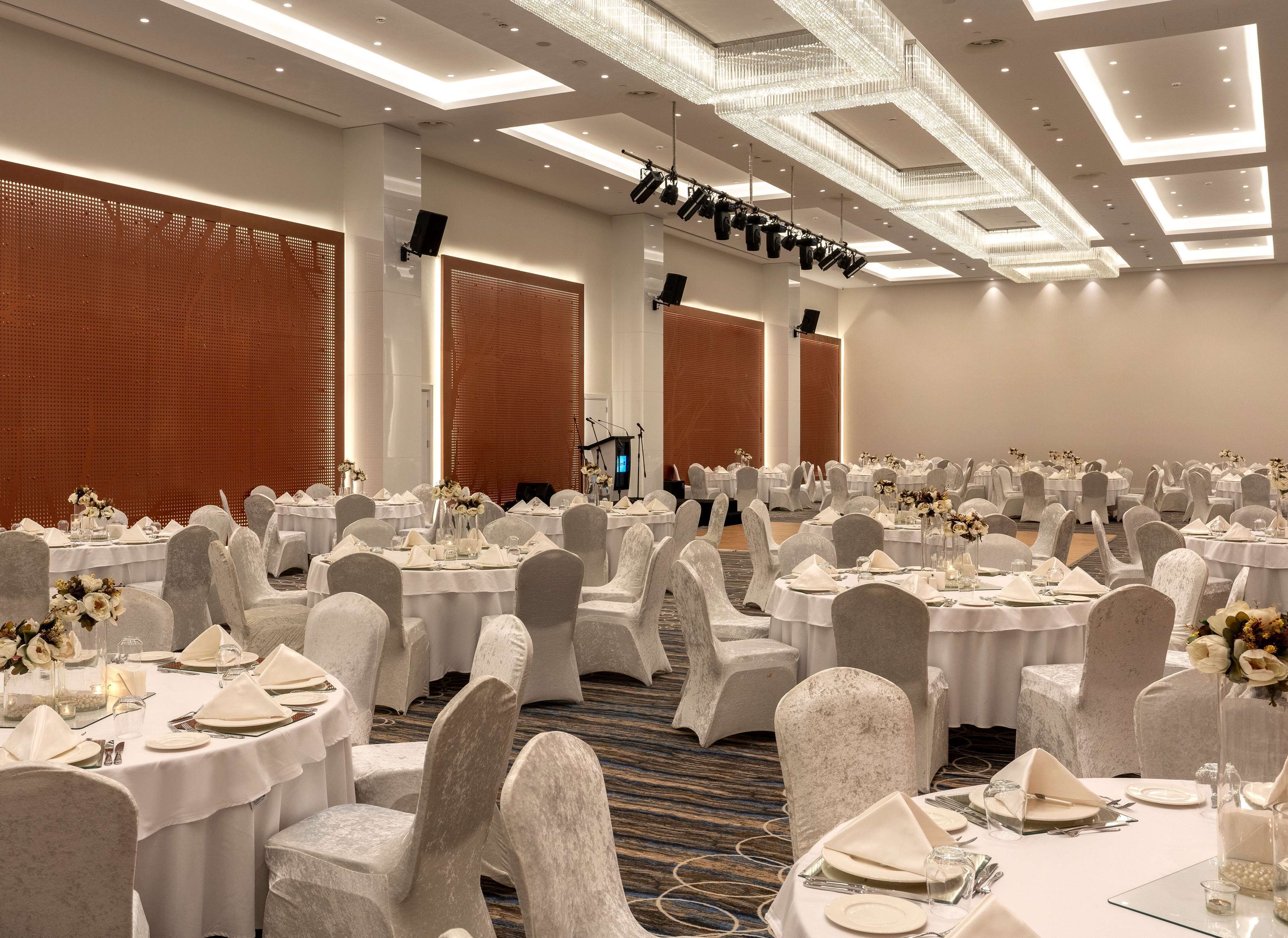 Uma sala de banquetes com mesas redondas, toalhas brancas e cadeiras cobertas por tecido branco.