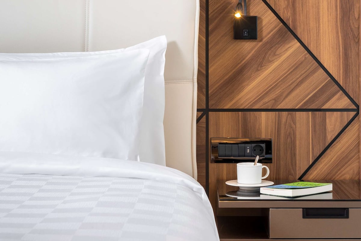 Quarto de hotel moderno com design de parede de madeira geométrica, cabeceira estofada branca, mesa de cabeceira com xícara e livro, e luminária pendente.