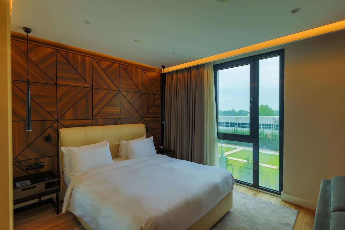 Quarto de hotel moderno com uma cama grande, uma parede de destaque em madeira, uma mesinha de cabeceira e vista para um jardim bem cuidado através de uma grande janela.