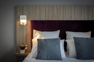 Cama bem arrumada com almofadas brancas e cinzentas, cabeceira escura e luminária fixada na parede sobre uma mesa de cabeceira de madeira com uma pequena planta.
