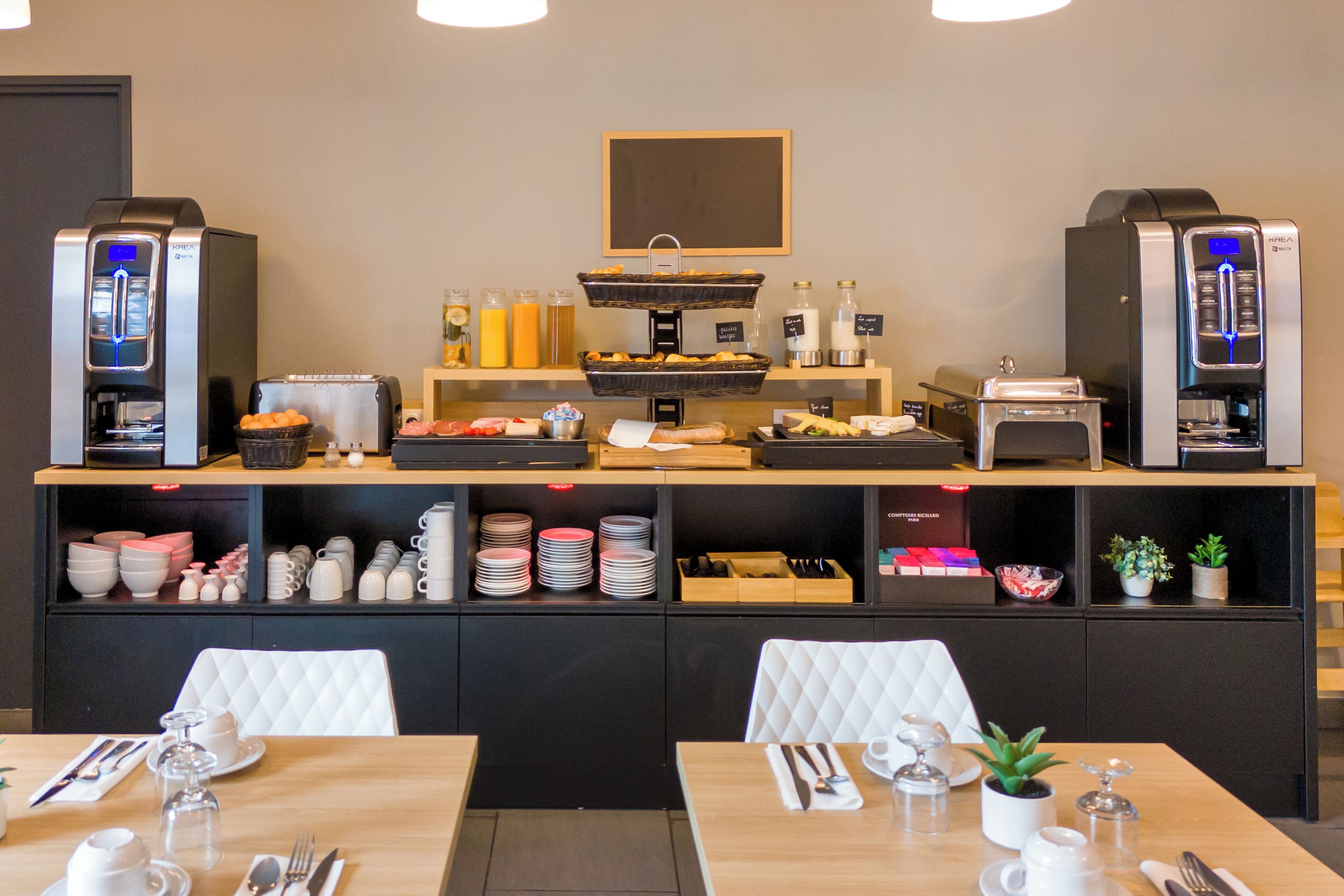 Espaço de buffet de café da manhã com máquinas de café expresso, diversas opções de comida e utensílios de mesa organizados.