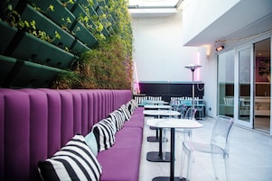 Uma área de estar moderna ao ar livre, com bancos acolchoados em roxo e cadeiras de plástico transparente, rodeada por vegetação e uma porta de vidro que dá para um espaço interior.