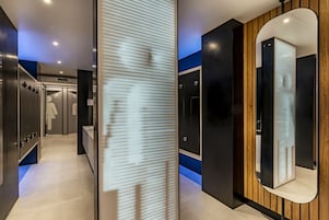 Corredor moderno de hotel com porta de vidro, parede de painéis de madeira e espelho refletindo o interior.