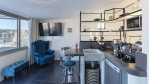 Uma cozinha moderna com fogão embutido, máquina de café e micro-ondas. Há um banco de bar e uma cadeira azul na sala de estar.