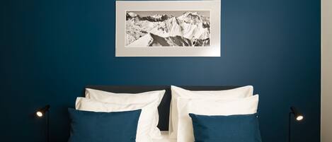 Quarto com cama, almofadas azuis e brancas, um quadro na parede e duas luminárias de cabeceira.