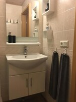 Casa de banho com lavatório branco, duas barras de toalhas, espelho e uma pequena estante com produtos de higiene pessoal.