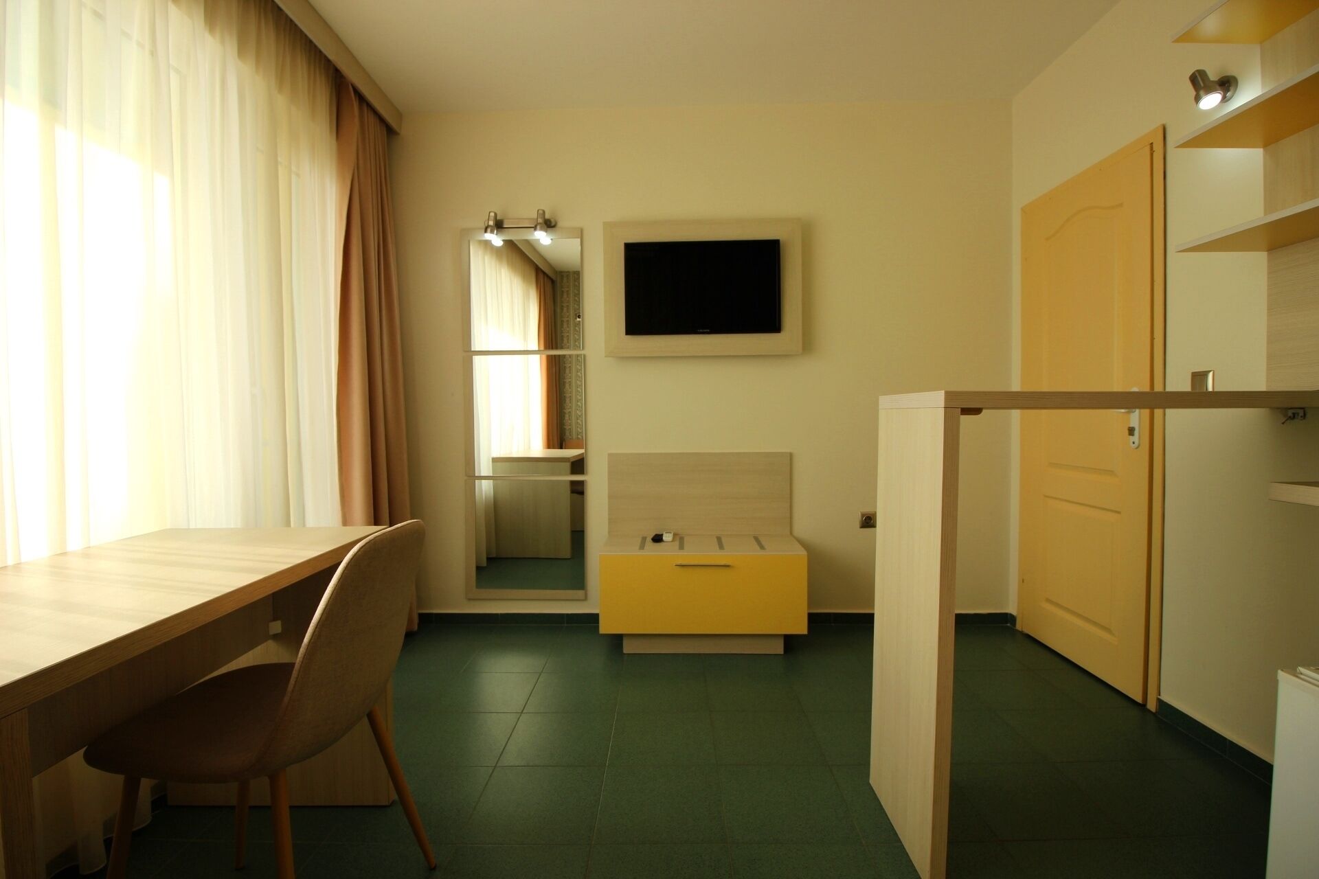 Quarto com escrivaninha de madeira, uma cadeira, cama com cabeceira amarela, televisão na parede e guarda-roupa.