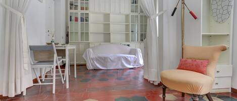 Quarto com escrivaninha branca, cama coberta, cadeira, estante e piso estampado.