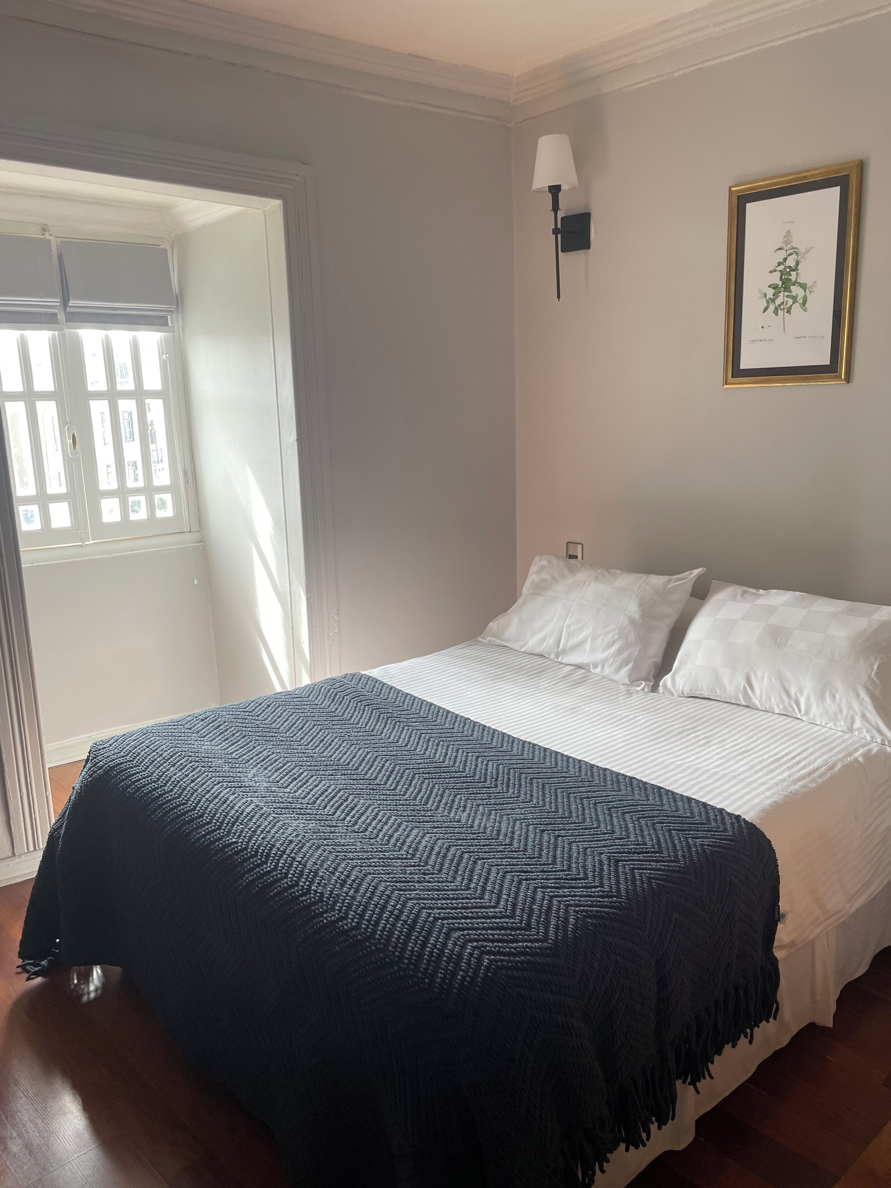 Quarto com cama, janela, luminária fixada na parede e ilustração botânica emoldurada.
