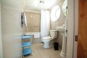 Casa de banho com sanita, lavatório e chuveiro. Há um carrinho azul e um porta-toalhas branco.